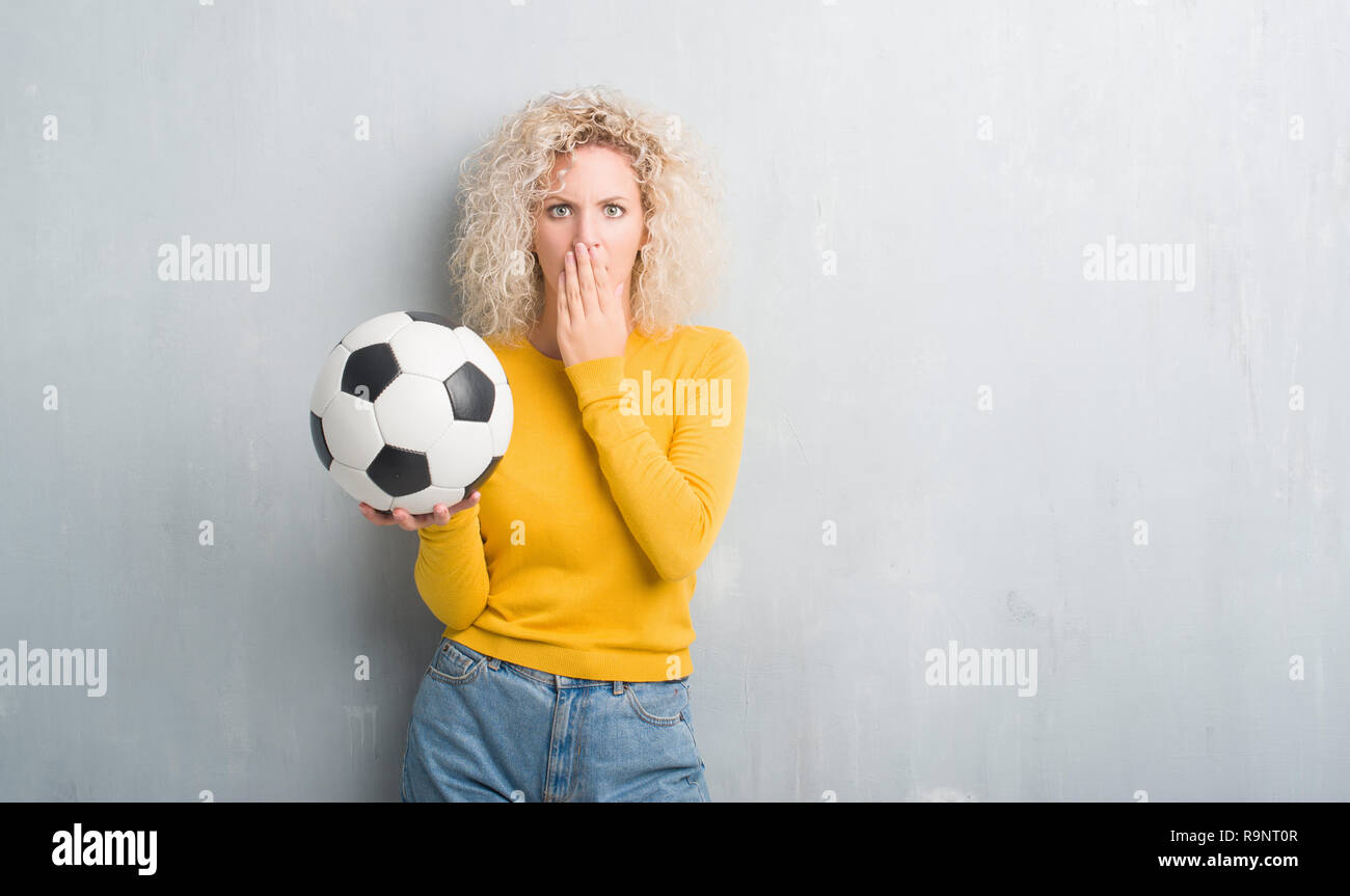 Young blonde woman over grunge grey background holding soccer football ...