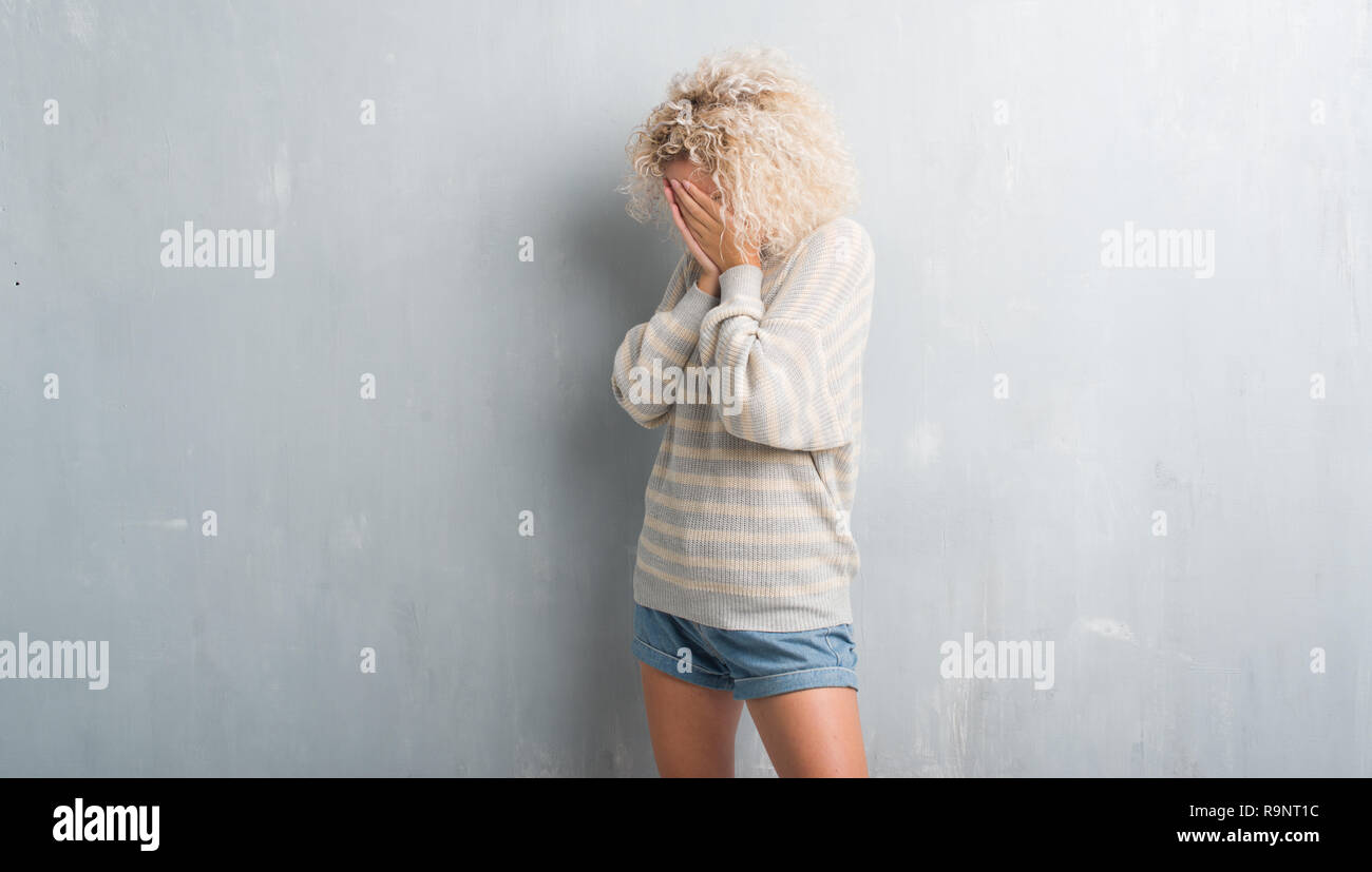 Young blonde woman with curly hair over grunge grey background with sad ...