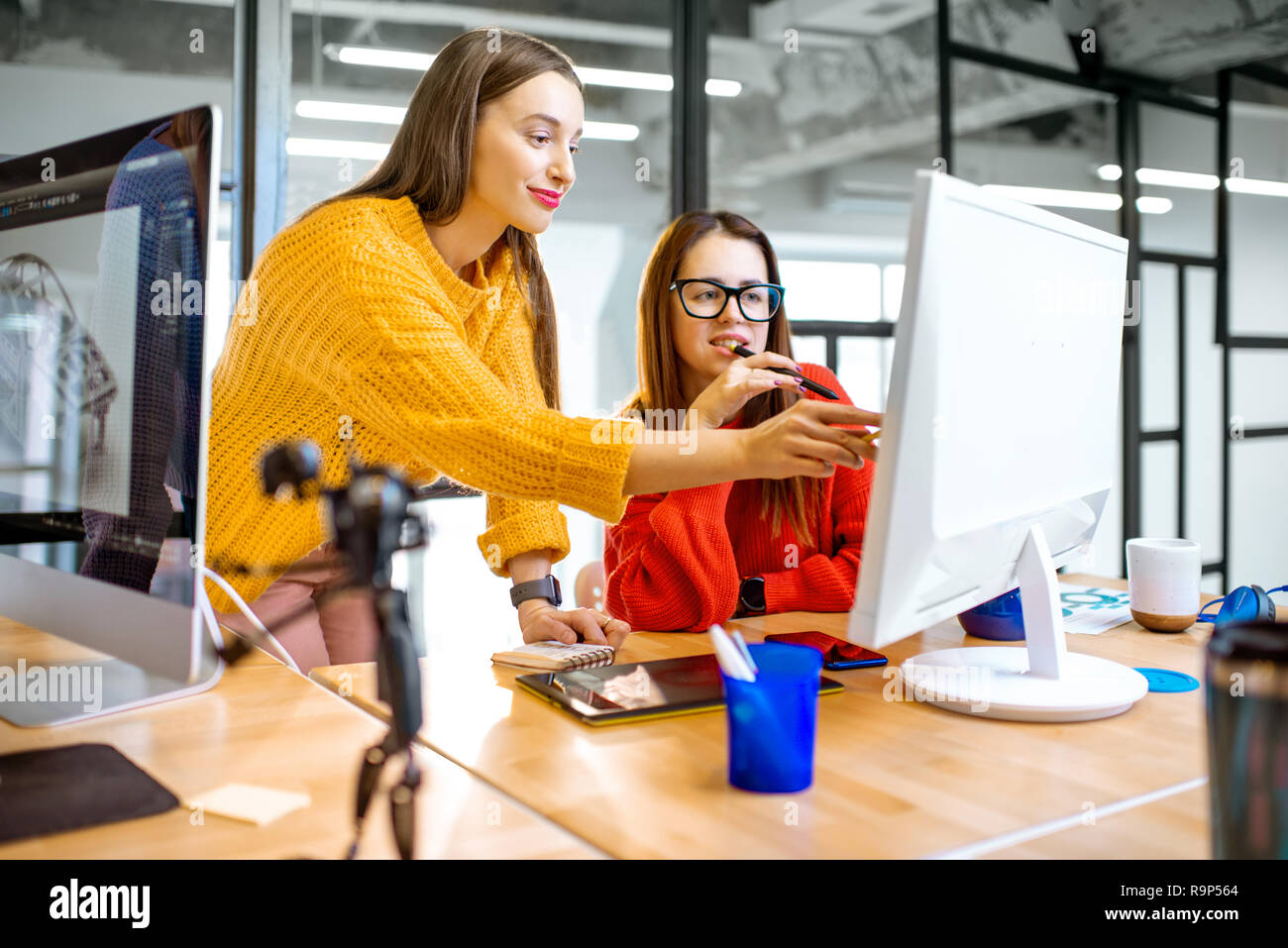 Two young creative women dressed in sweaters working with computer in ...