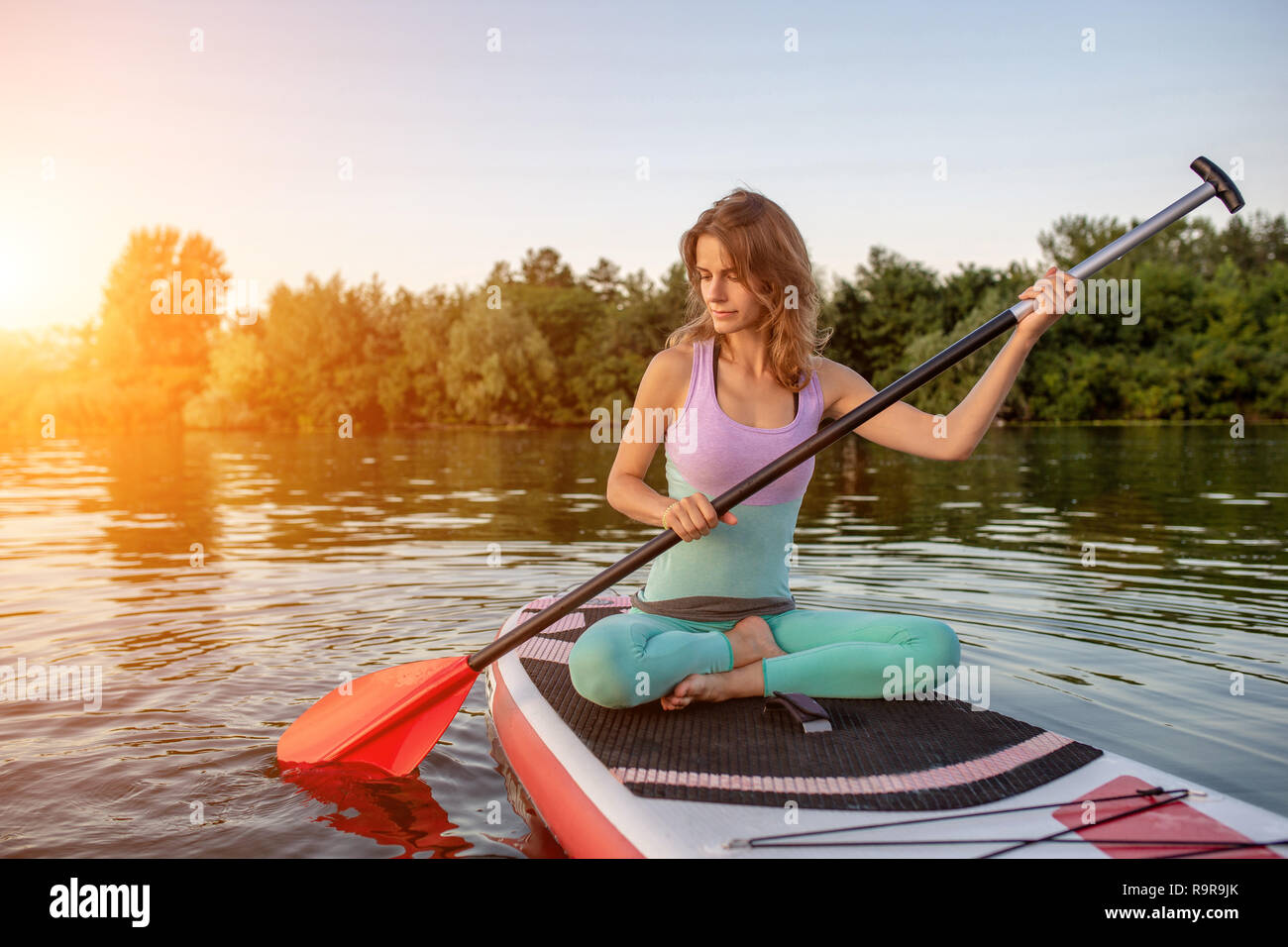 Young woman sitting on paddle board, practicing yoga pose. Doing yoga ...