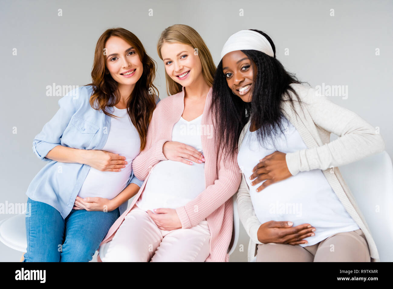 multicultural pregnant women sitting close together, touching bellies ...