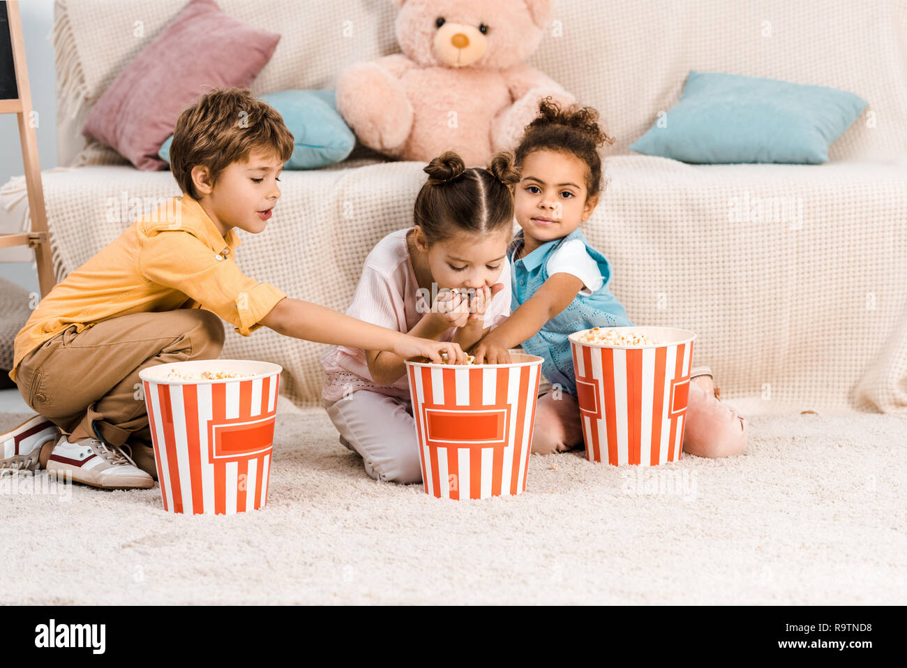 cute multiethnic children sitting on carpet and eating popcorn Stock ...