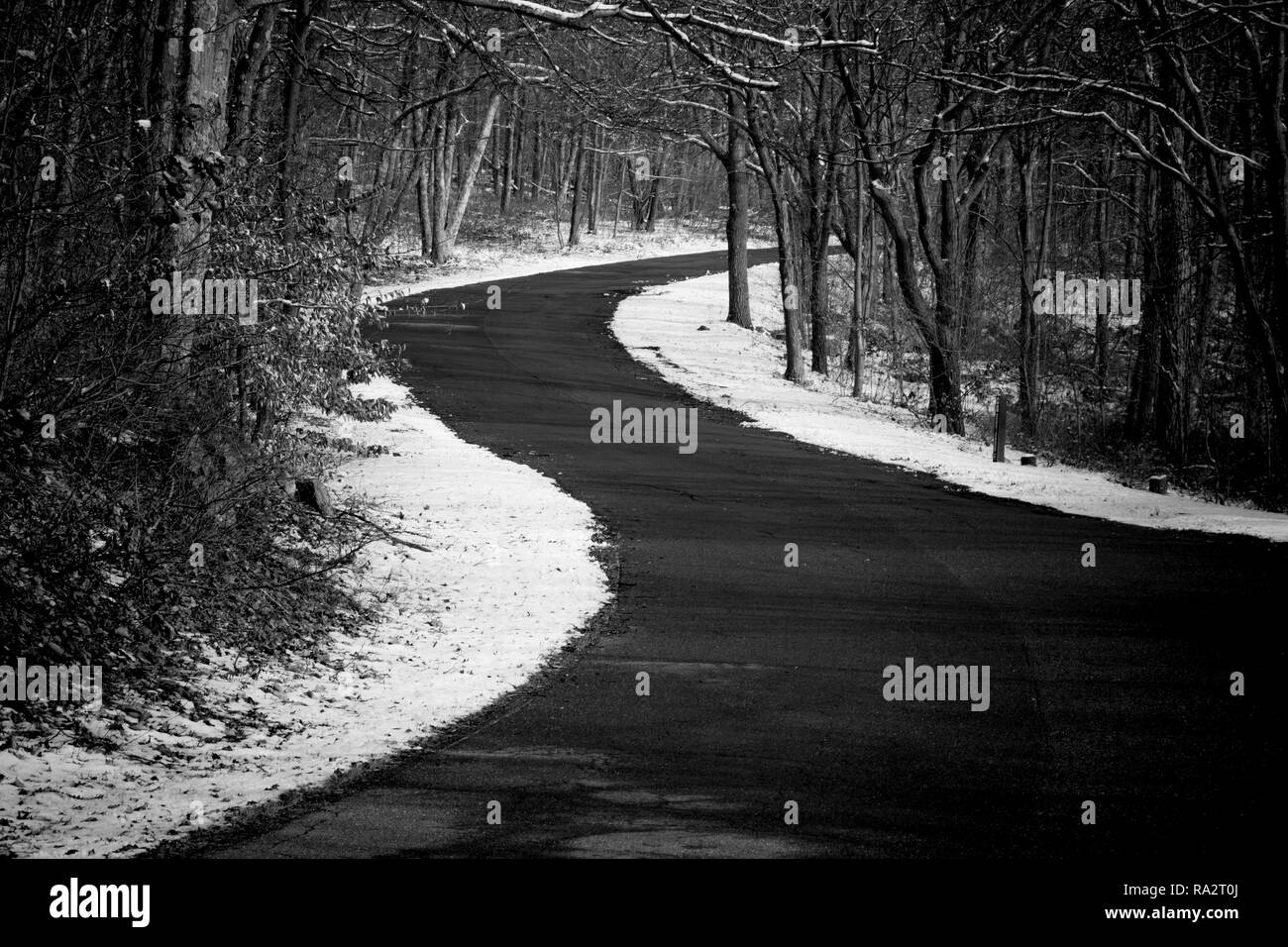 A country road curves uphill surrounded by snow in winter, black and ...