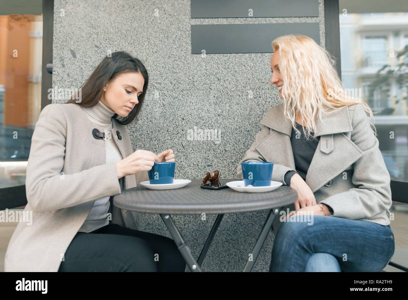 Two young smiling women in an outdoor cafe, drinking coffee, talking ...