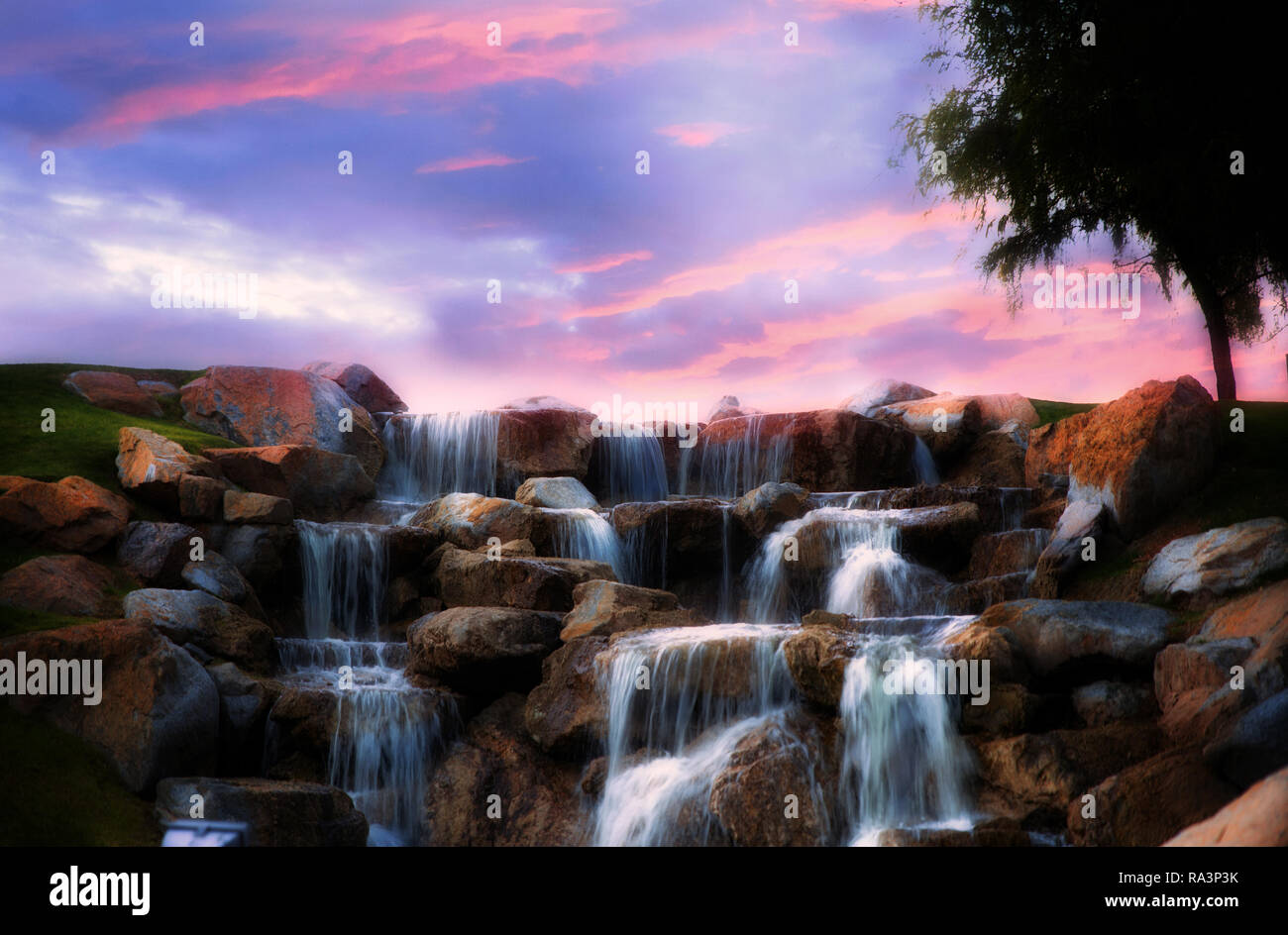 Water falling softly over large boulders between green banks of grass ...