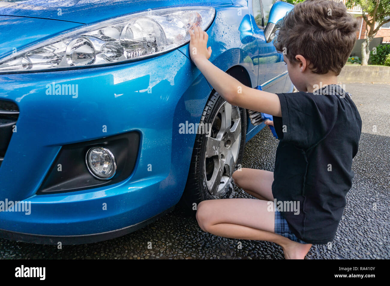 Boy earning pocket money cleaning blue compact car with hose, bucket of ...