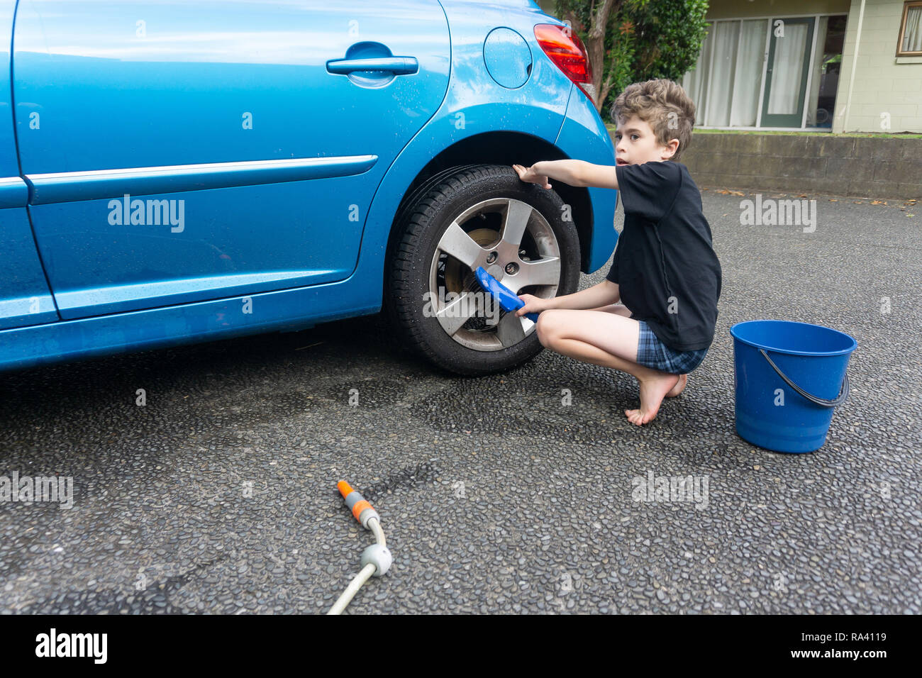 Boy earning pocket money cleaning blue compact car with hose, bucket of ...