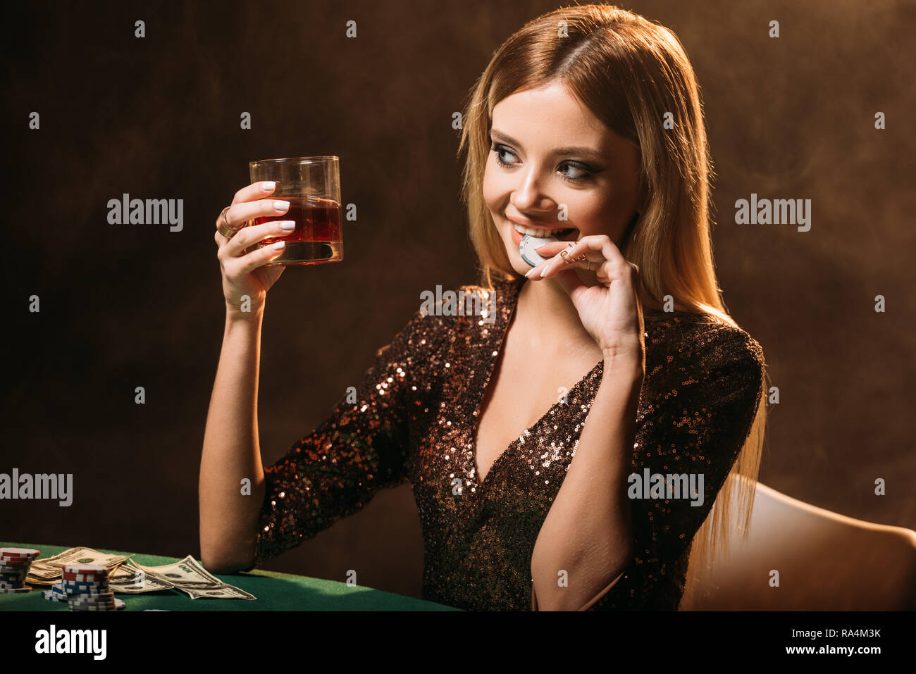 attractive smiling girl holding glass of whiskey and biting poker chip ...