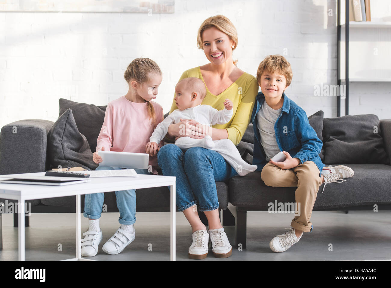 happy mother smiling at camera while sitting on couch with three ...