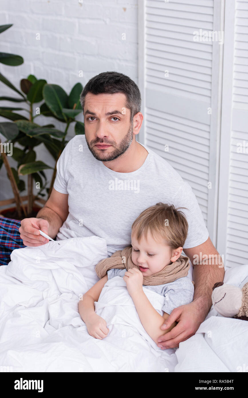 dad hugging sick son in bedroom and checking his temperature with ...