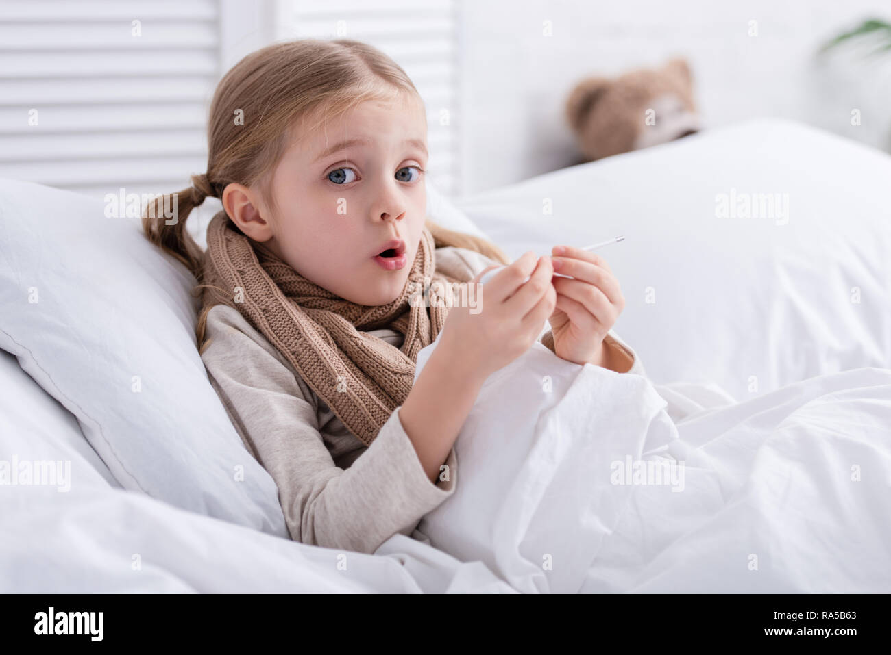shocked sick kid with scarf over neck lying in bed and checking temperature with thermometer at ...