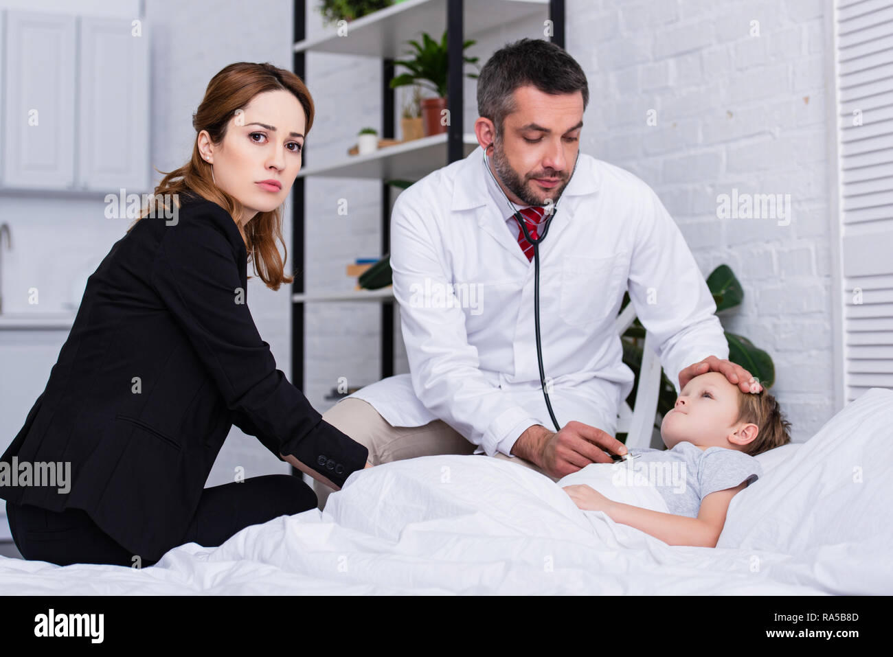 pediatrist in white coat examining sick boy with stethoscope, mother in ...