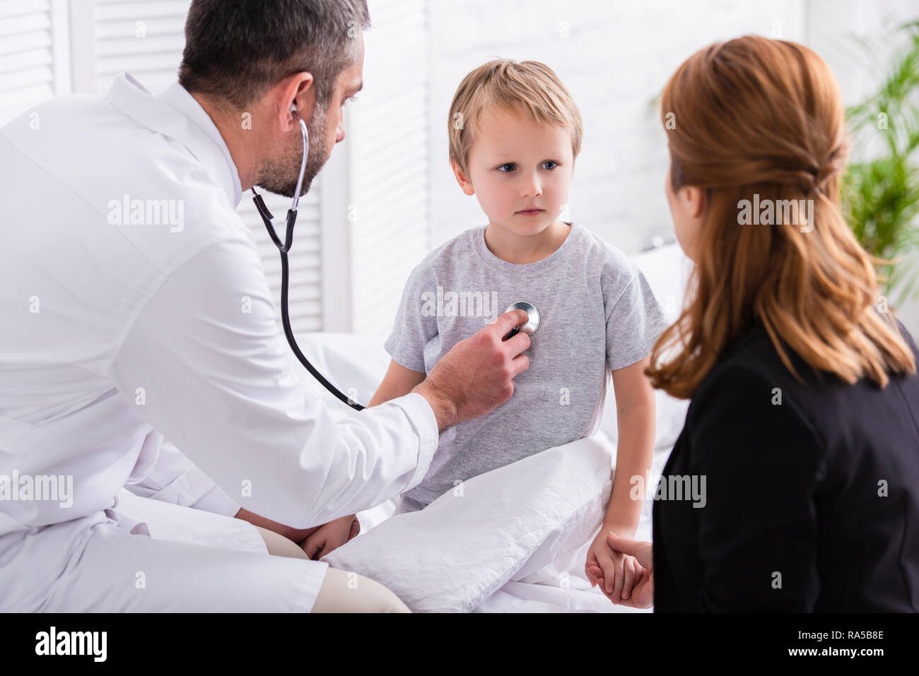 pediatrist in white coat examining sick boy with stethoscope at home ...