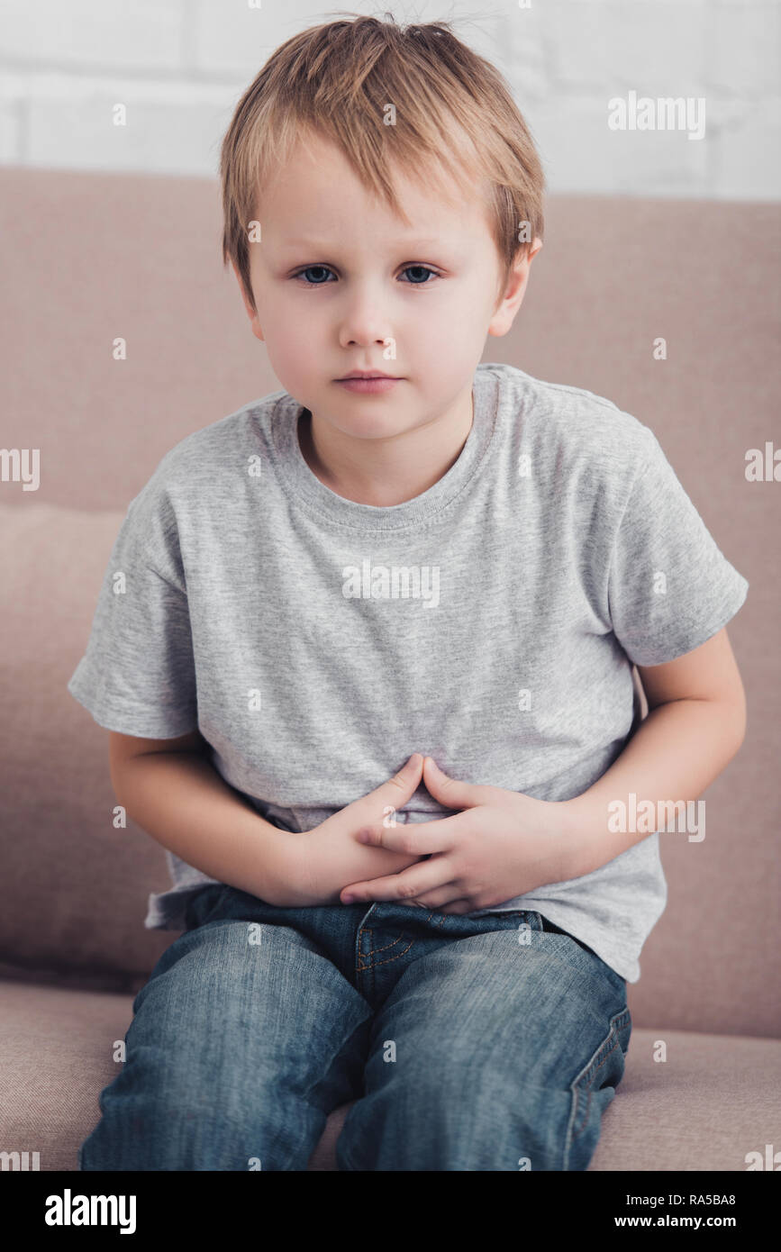 sick boy with stomach pain sitting on sofa in living room and looking ...