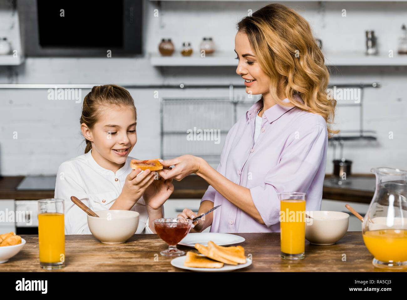 smiling woman giving toast with jam to cute daughter at table in ...