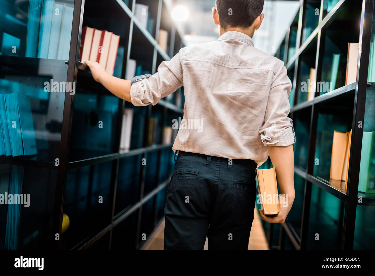 back view of boy holding book and walking between bookshelves in ...