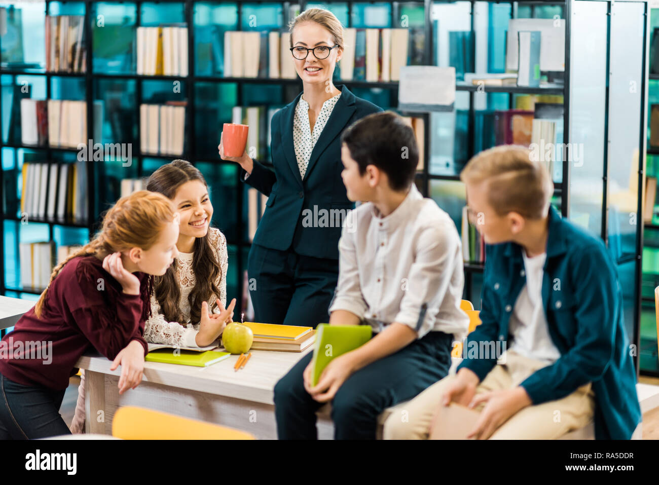 young female librarian holding cup and looking at smiling schoolkids in ...