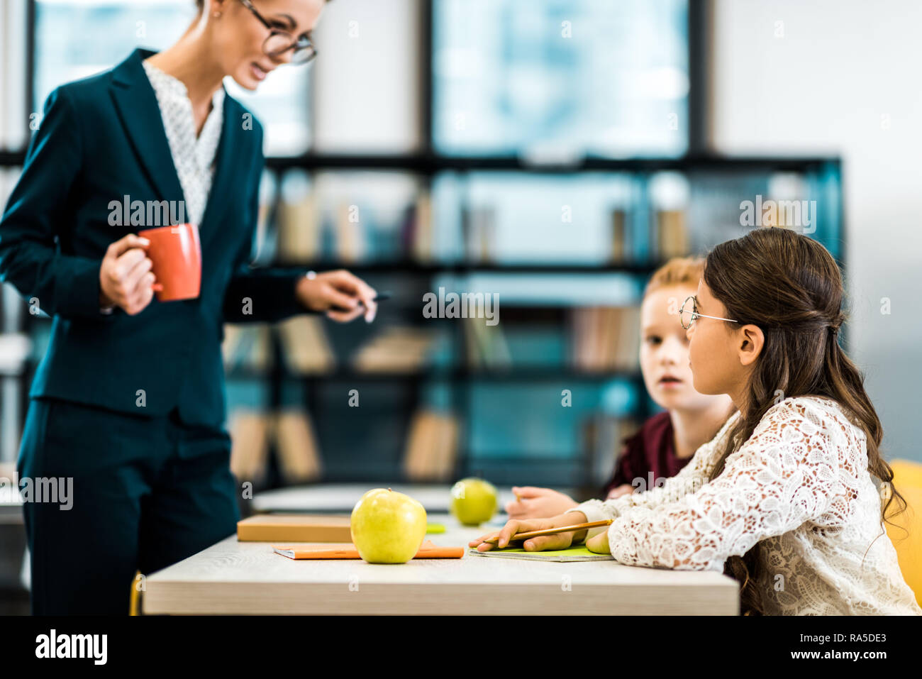 young teacher holding cup and looking at schoolkids studying in library ...