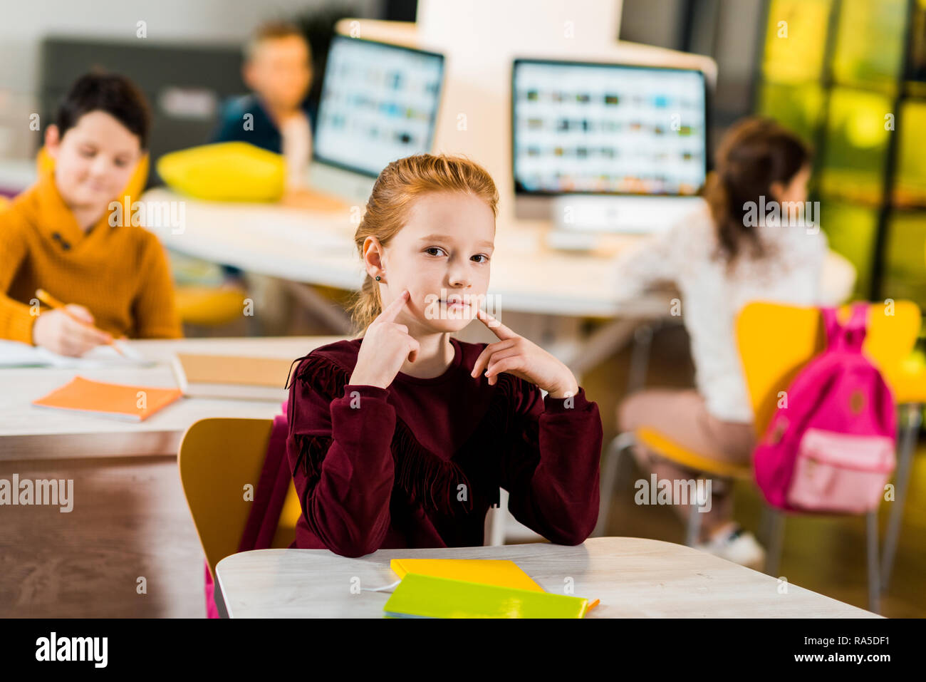 adorable schoolkid looking at camera while sitting with classmates in ...