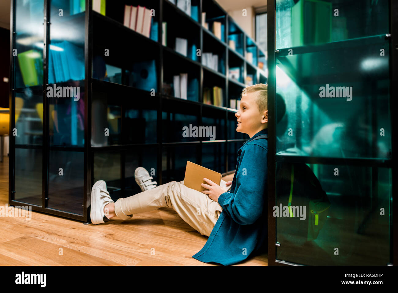 side view of smiling boy sitting on floor and reading book in library ...