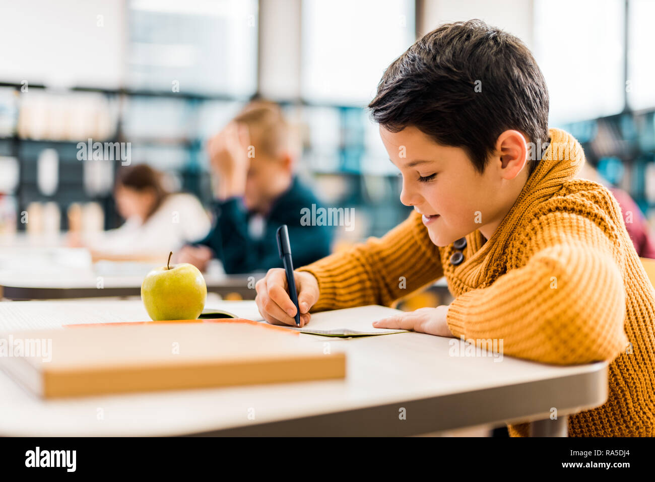 smiling boy writing with pen while studying with classmates in library ...