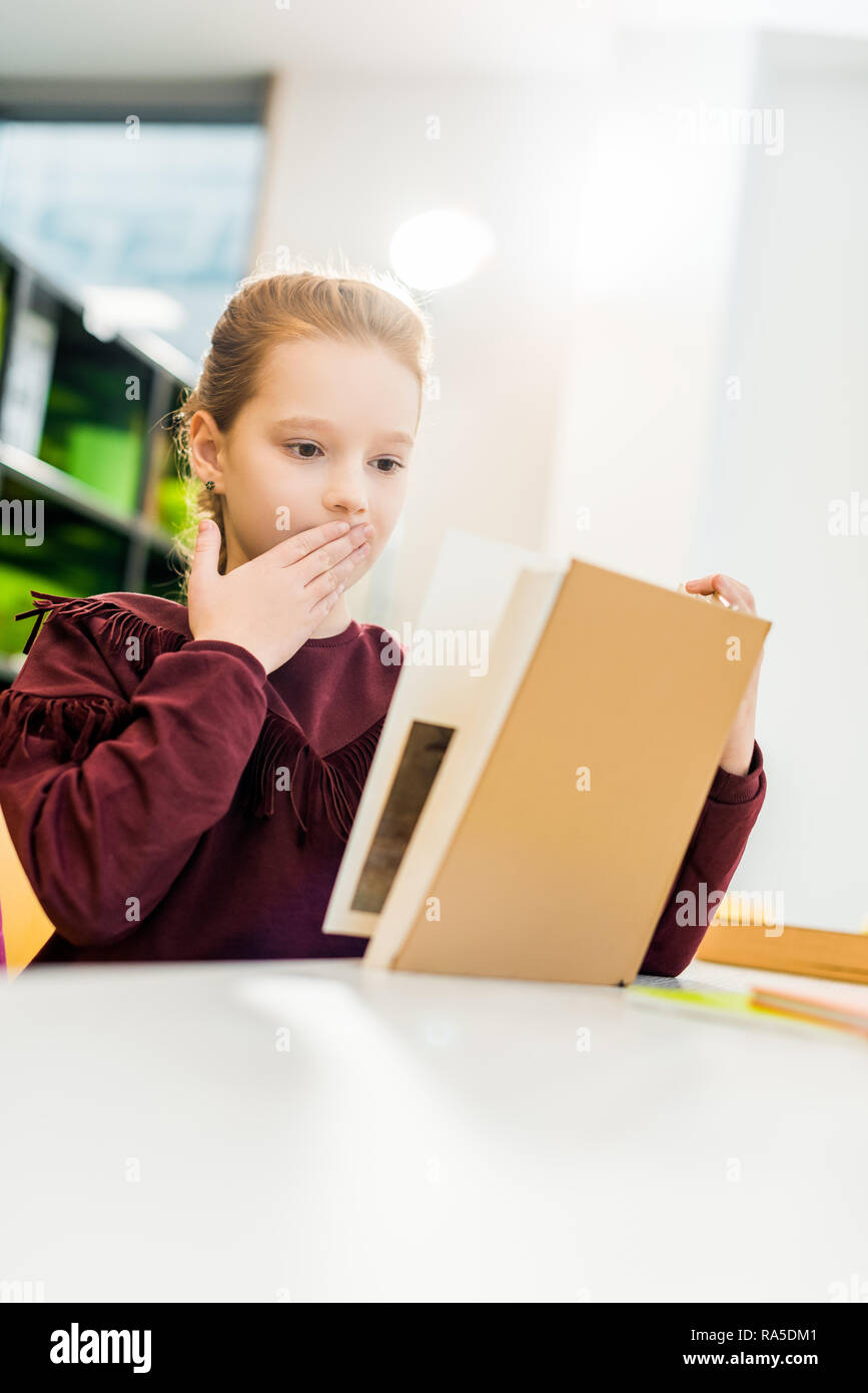 shocked schoolgirl reading book while sitting in library Stock Photo ...