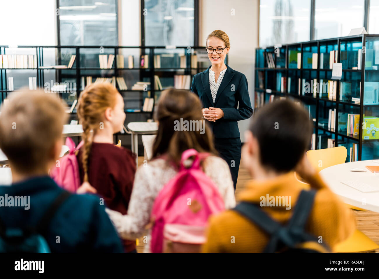back view of schoolkids with backpacks looking at female librarian ...
