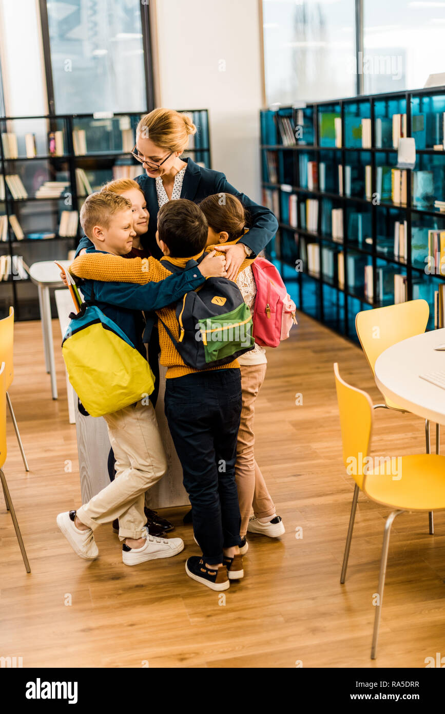 happy kids with backpacks hugging librarian in library Stock Photo - Alamy