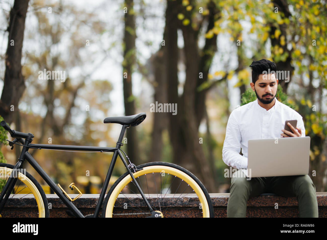 Happy young indian man using mobile phone while working on laptop ...