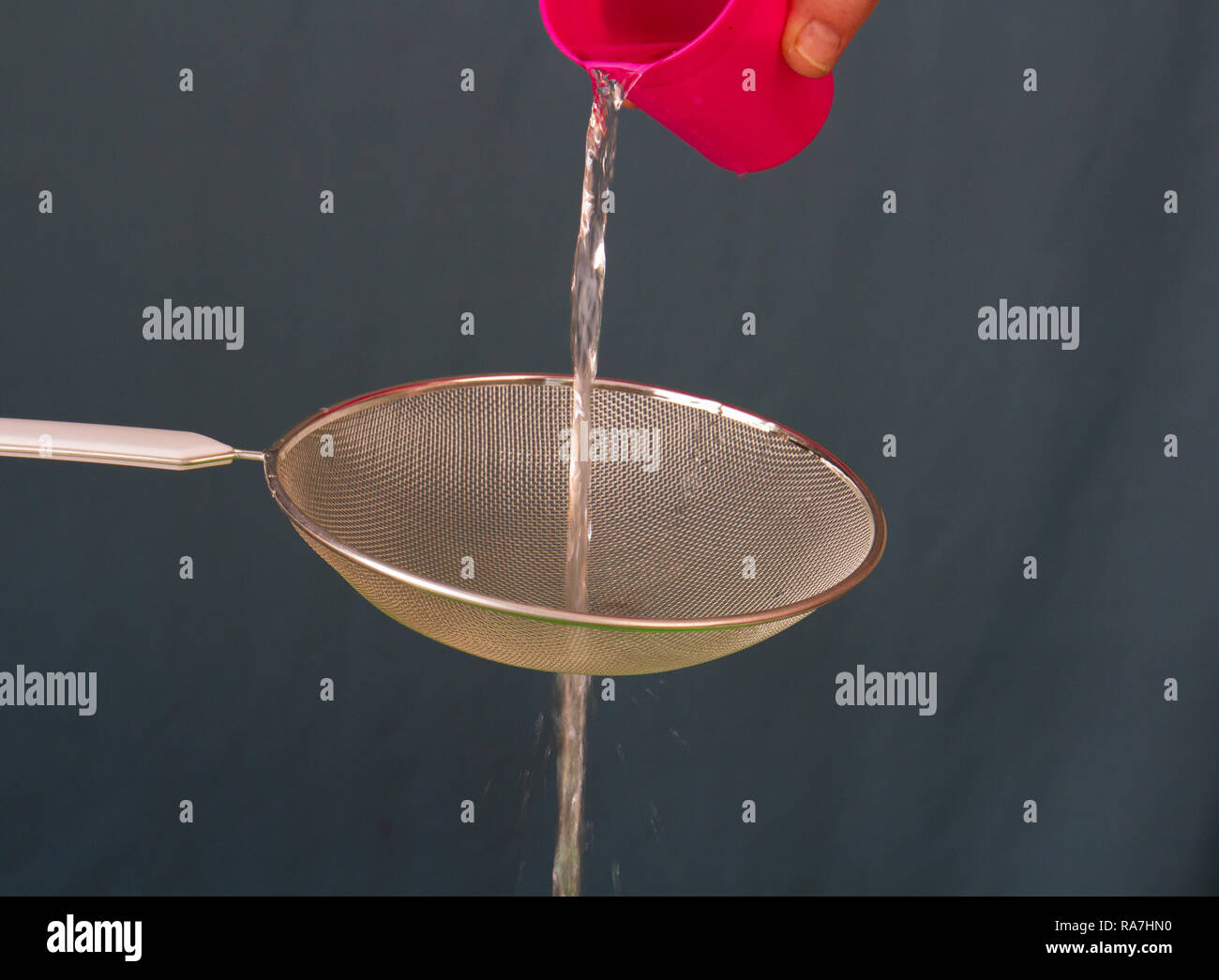 Water poured from a pink mug runs through a sieve isolated on a clear ...