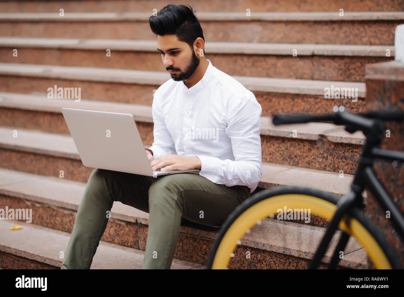 Young indian student working on laptop while sitting on stairs at the ...