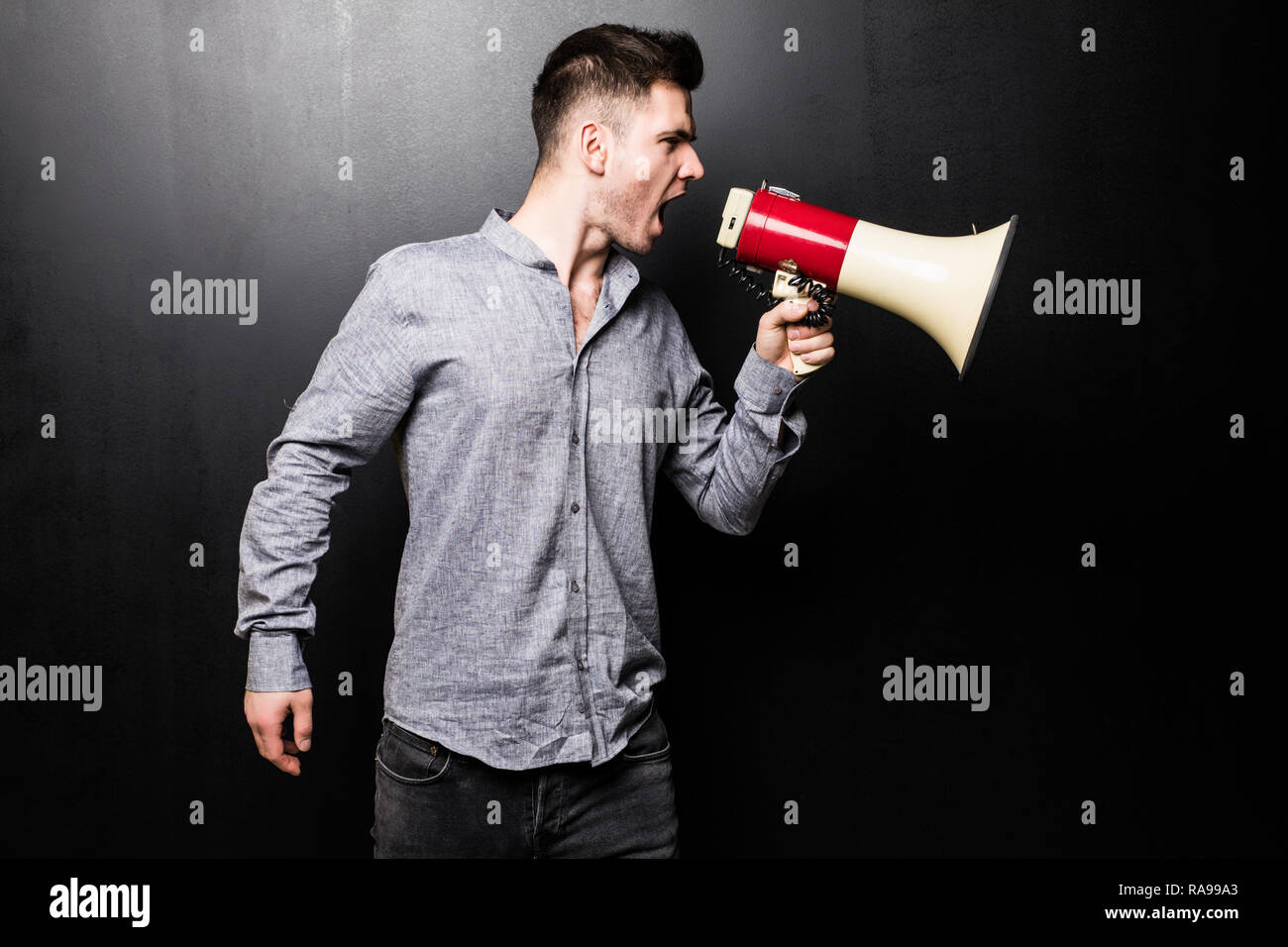 Portrait of young man yelling into the megaphone over black background ...