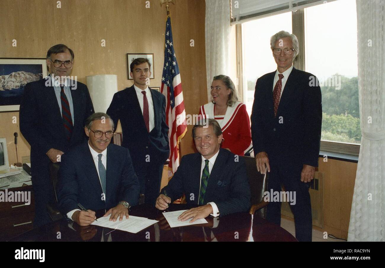 TWO MEN SIGNING PAPERS AT THE TABLE WITH THREE MEN AND A WOMEN STANDING ...