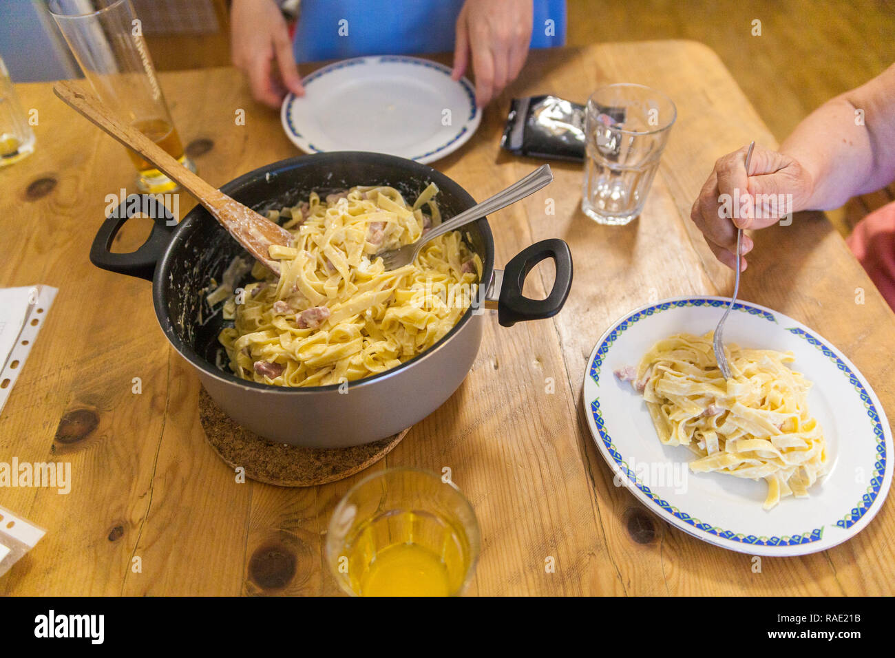 A pot full of pasta carbonara, two plates on a table. A young women is ...