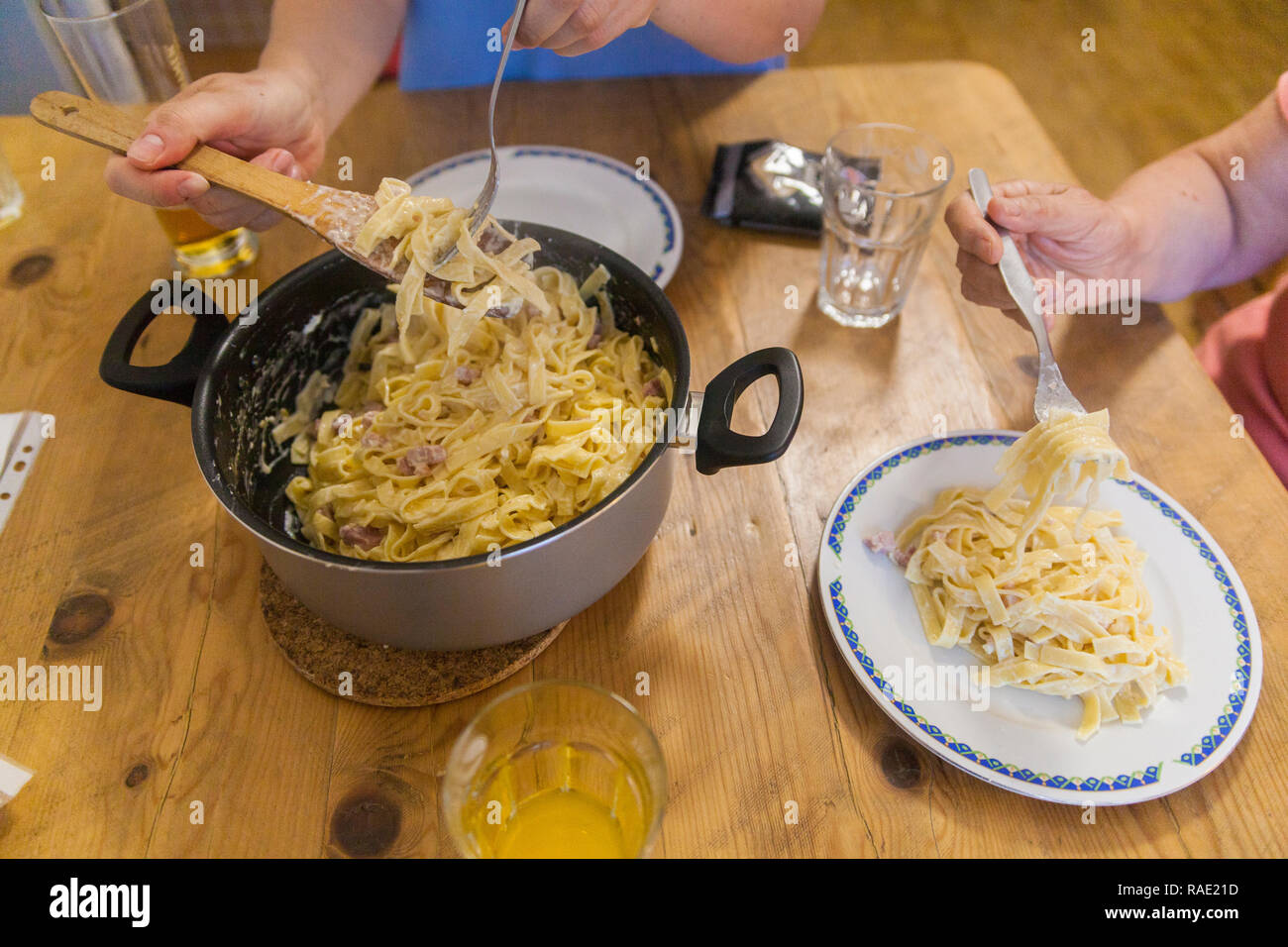 A pot full of pasta carbonara, two plates on a table. A young women is ...