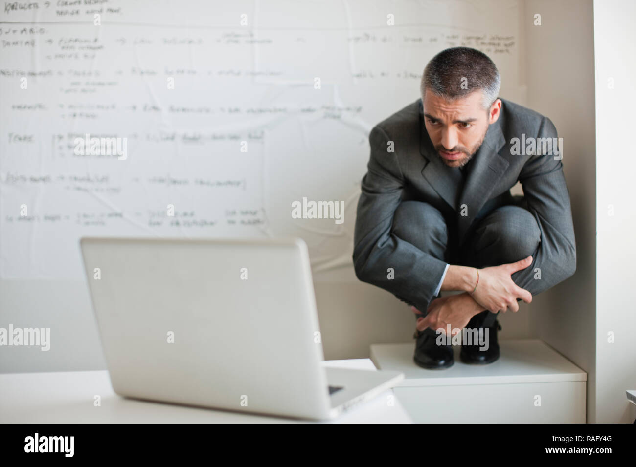 Businessman squats on desk looking at a laptop screen seemingly ...