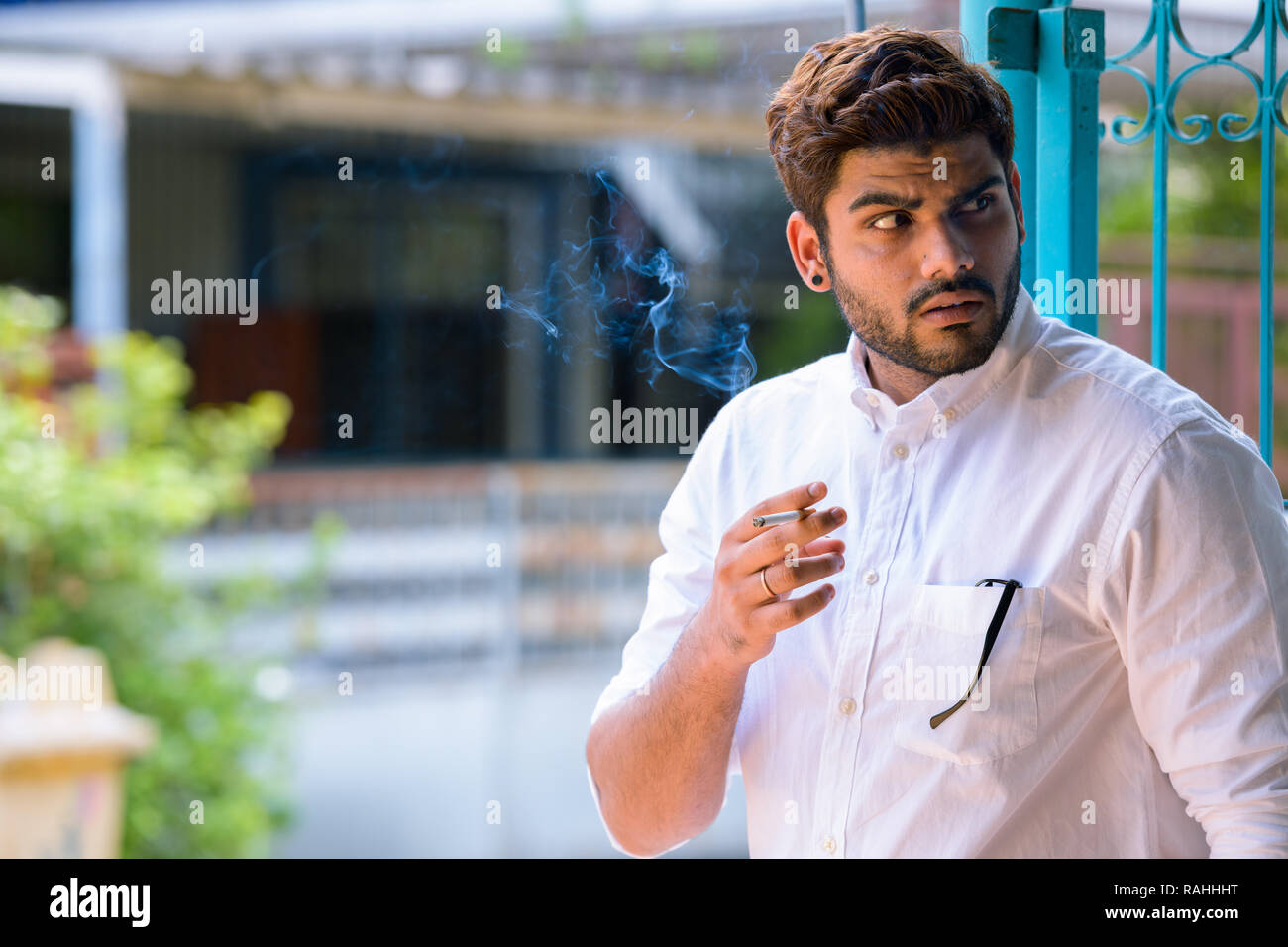Young bearded Indian man thinking while smoking cigarette outdoors ...