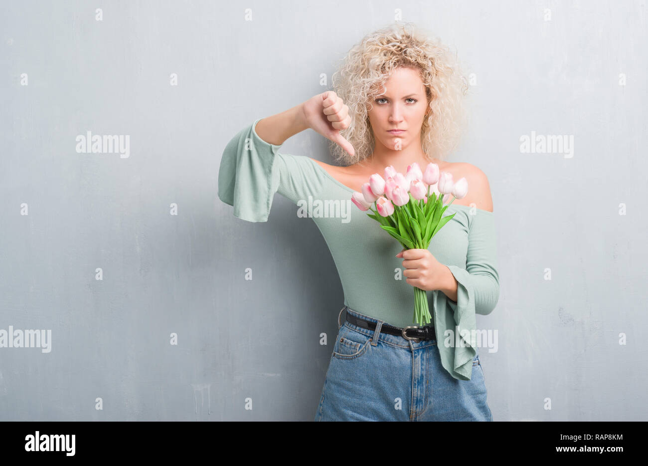 Young blonde woman over grunge grey background holding flowers with ...