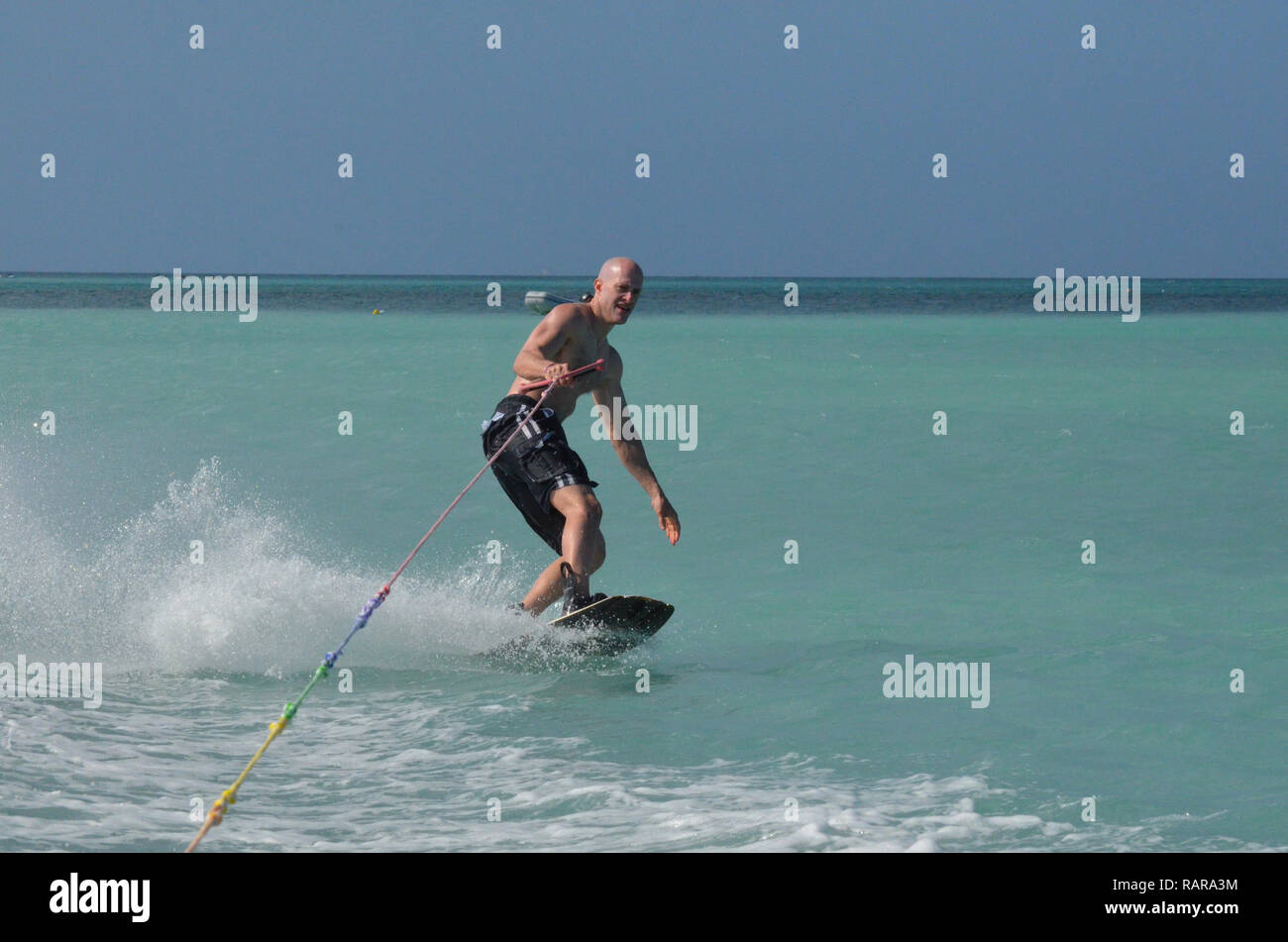 Bald guy wakeboarding with water spraying around his feet in Aruba ...