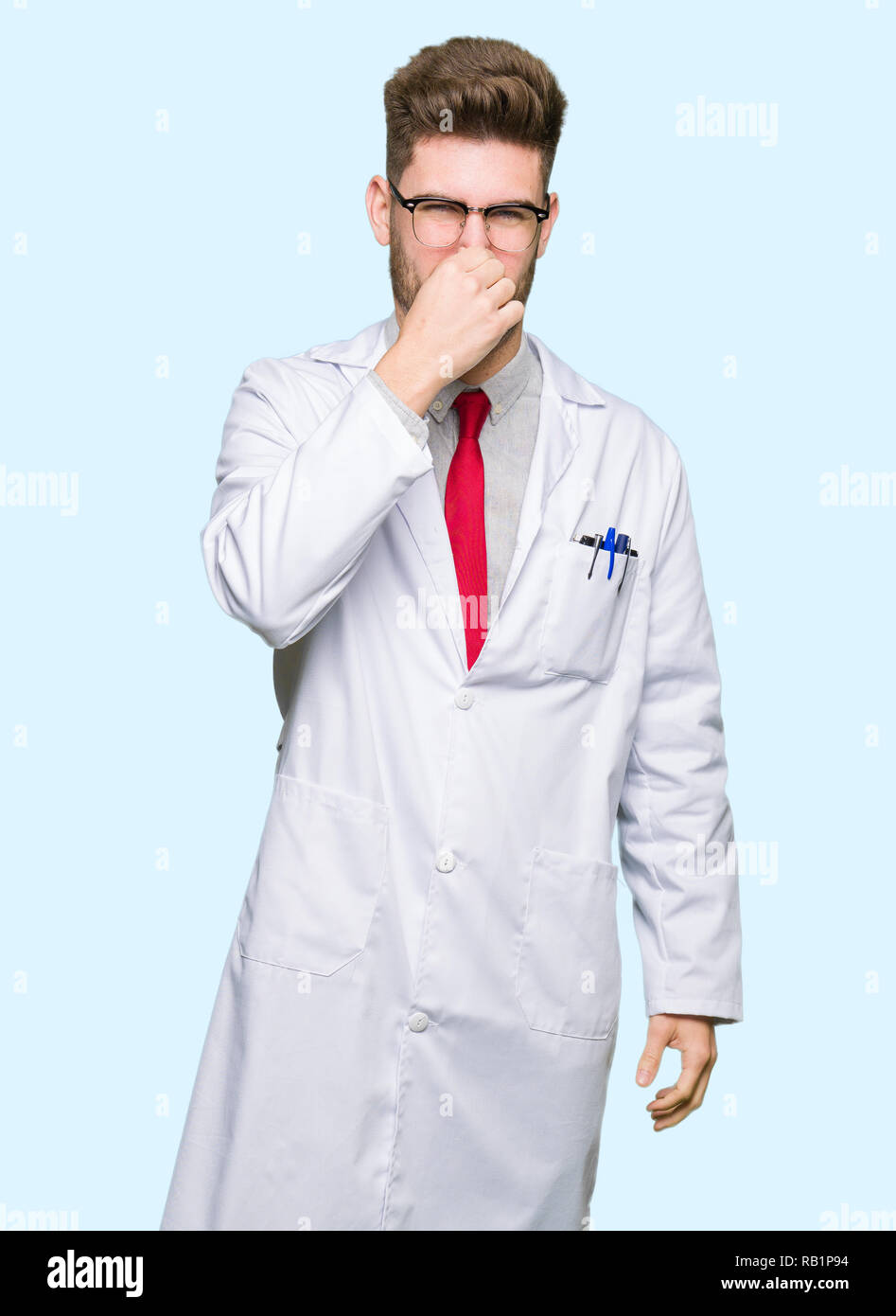 Young handsome scientist man wearing glasses smelling something stinky ...