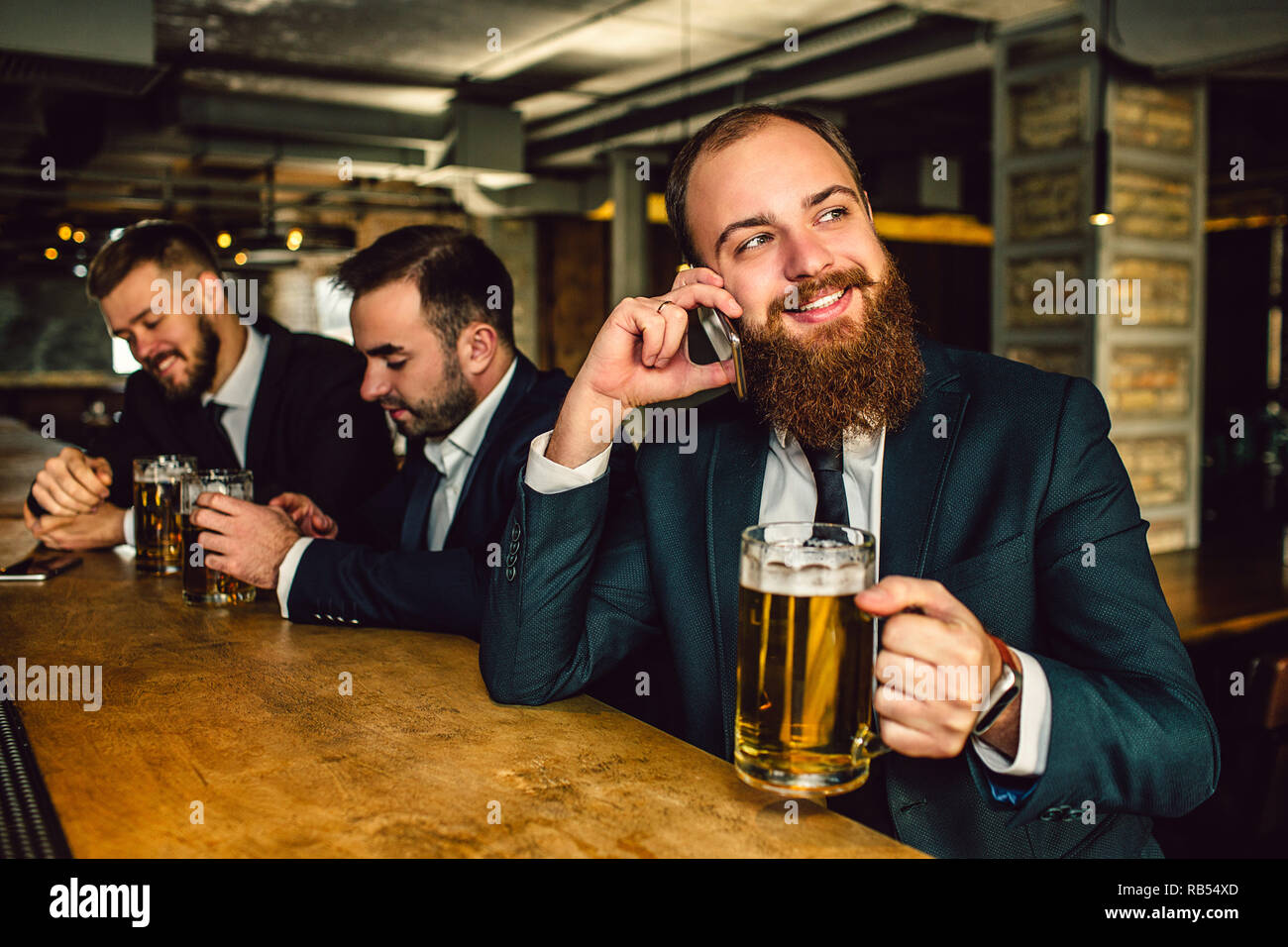 Young bearded man in suit talk on phone. He sit at bar counter and hold ...