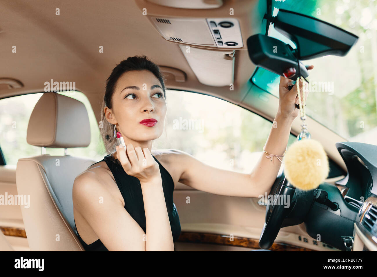 Young elegant woman looking in rear view mirror painting her lips doing ...