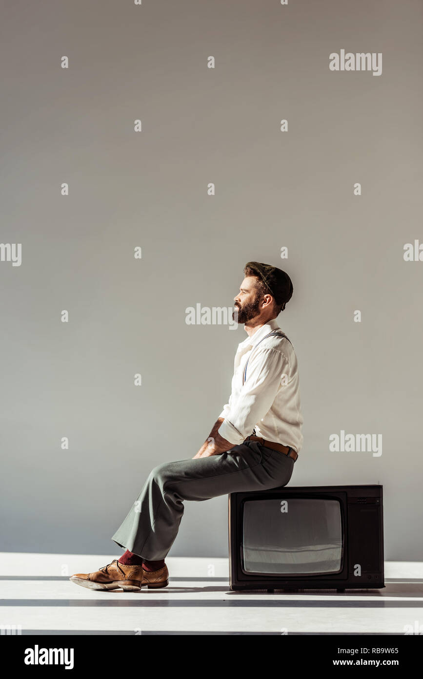 side view of handsome bearded man sitting on vintage tv on grey ...