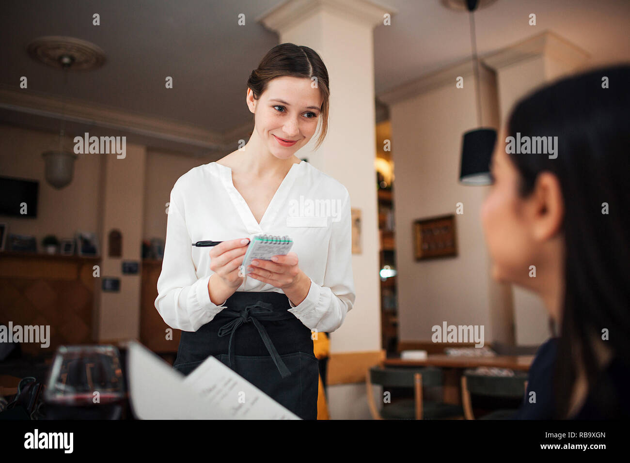 Cheerful young waitress stand in front of customer. She write order ...