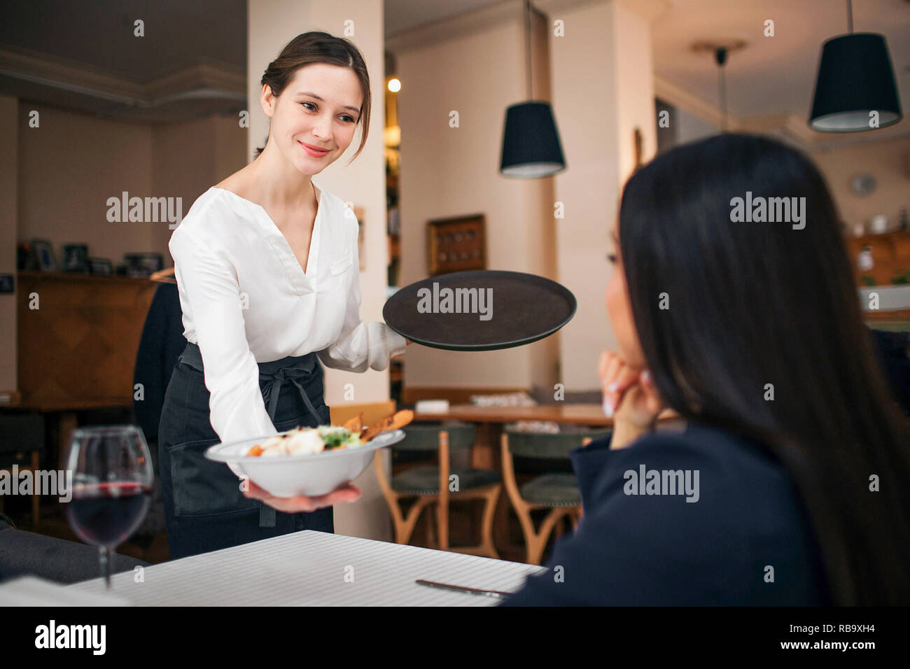 Pleasant young waitress stand at table with sitting customer and show ...
