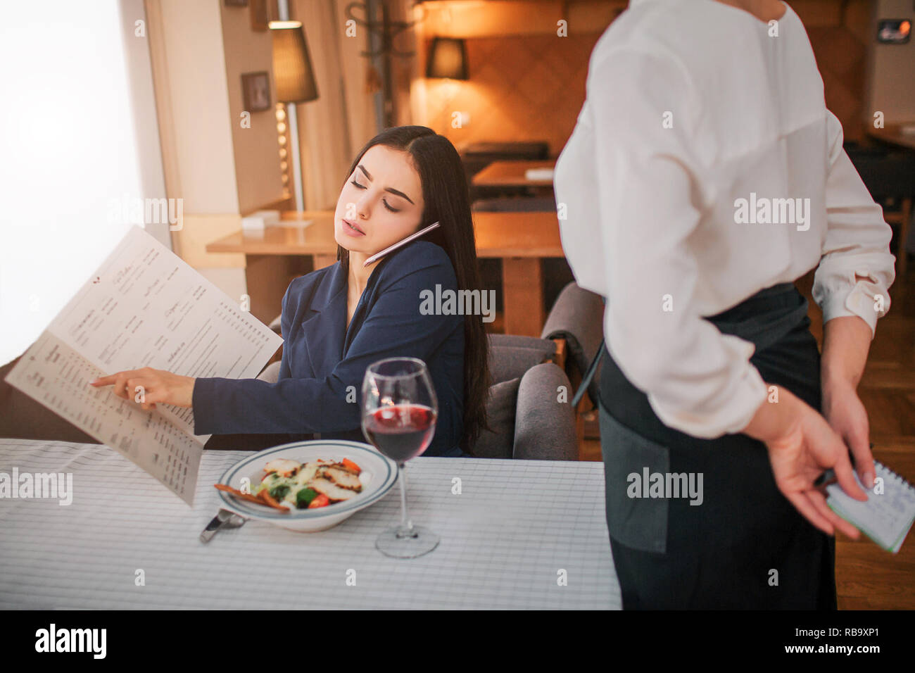 Busy young woman sit at table and talk on phone. She hold menu and ...