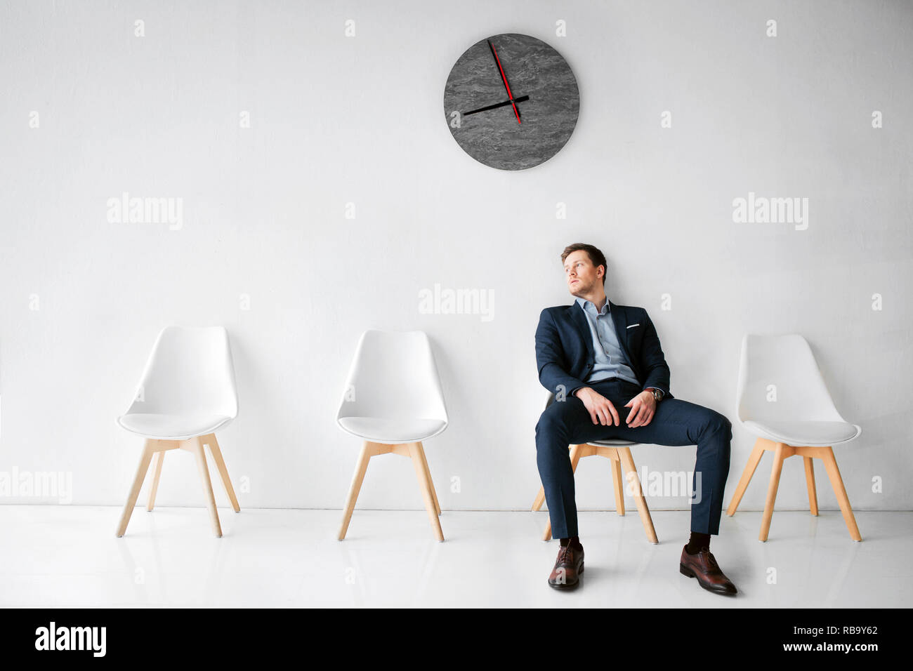 Young man in suit sit on white chair in white room. He lean to wall ...