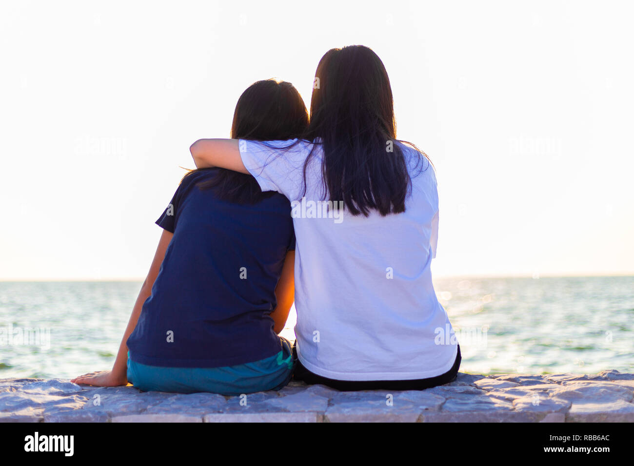 Two young sisters sitting on stone wall watching sunset over the ocean ...