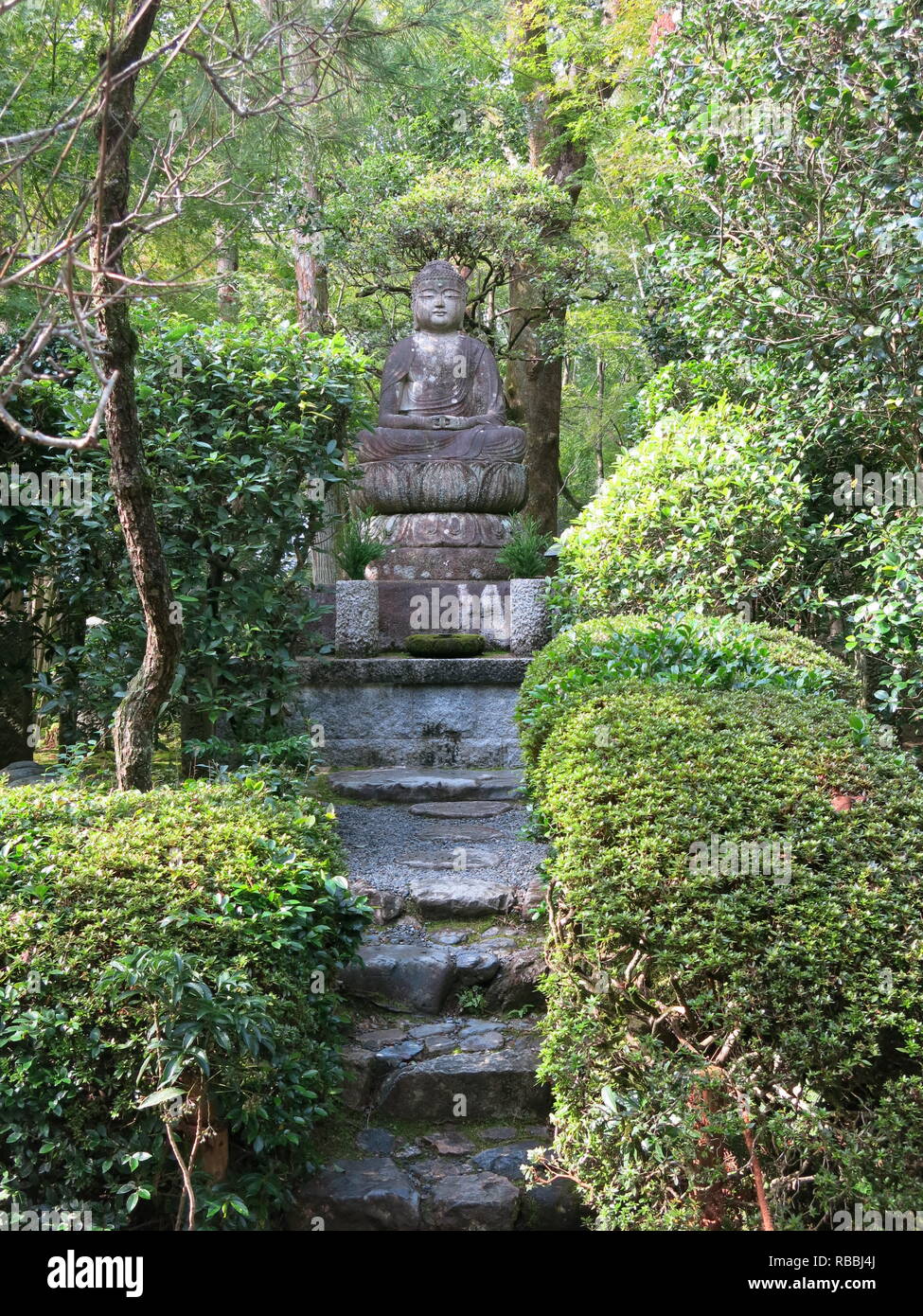 A tall stone statue with a Buddha on top, is flanked by hedges at the ...