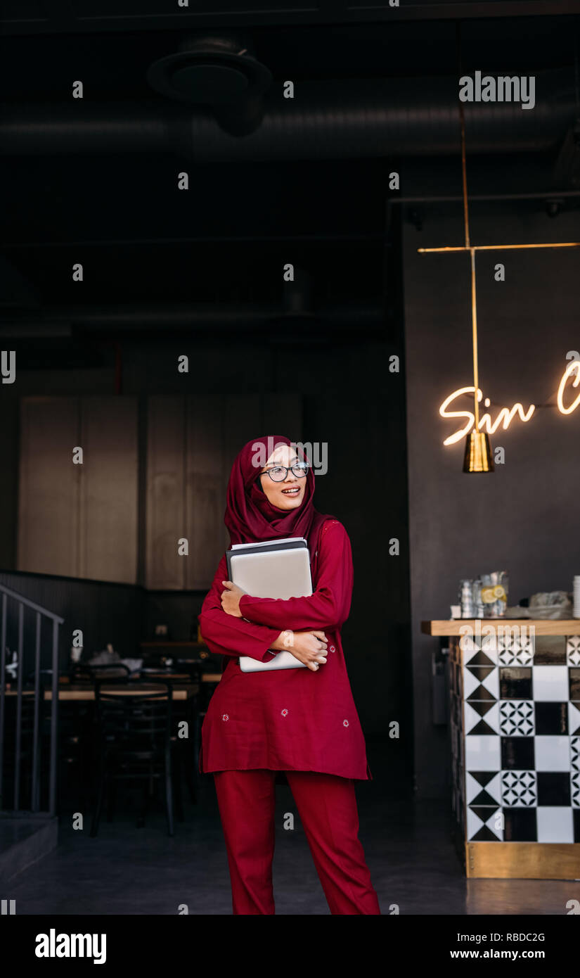 Woman in hijab grabbing laptop with both hands and looking away while ...